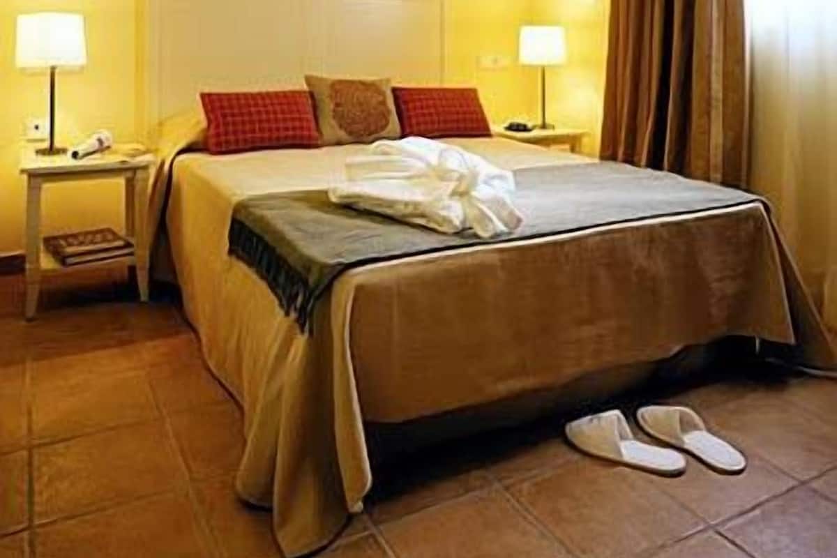 Quarto de hotel com cama, duas mesas de cabeceira, luminárias, um telefone e um par de chinelos no chão.