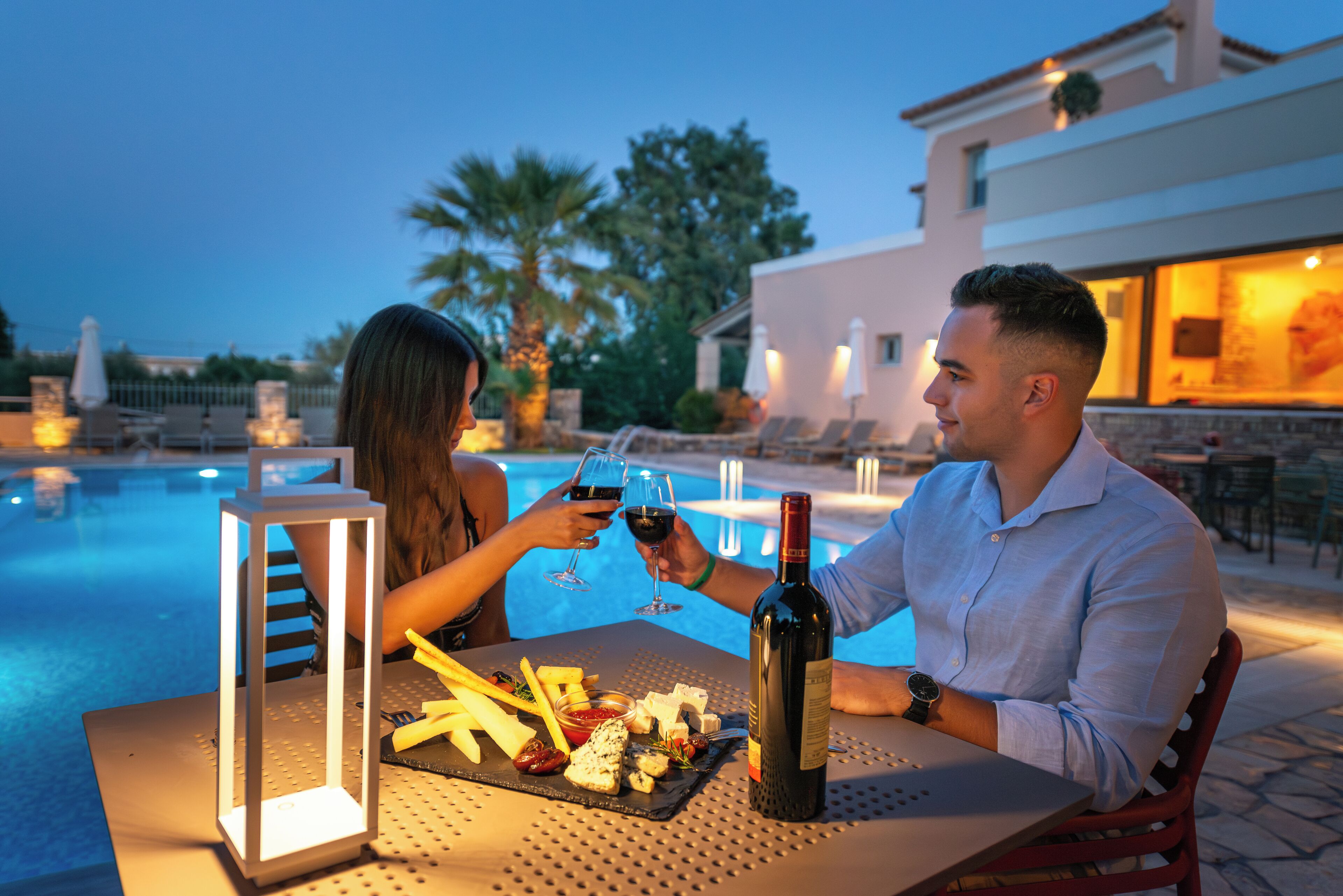 Um casal sentado à mesa perto da piscina, com uma garrafa de vinho, copos e um prato de comida.