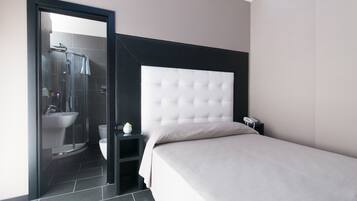 Quarto de hotel moderno com uma cama grande, uma mesinha de cabeceira com telefone e área de chuveiro com divisória de vidro.