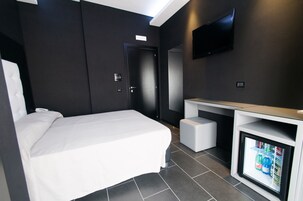 Quarto de hotel moderno com cama, um pequeno espaço de estar, televisão na parede e mini-geladeira com bebidas.