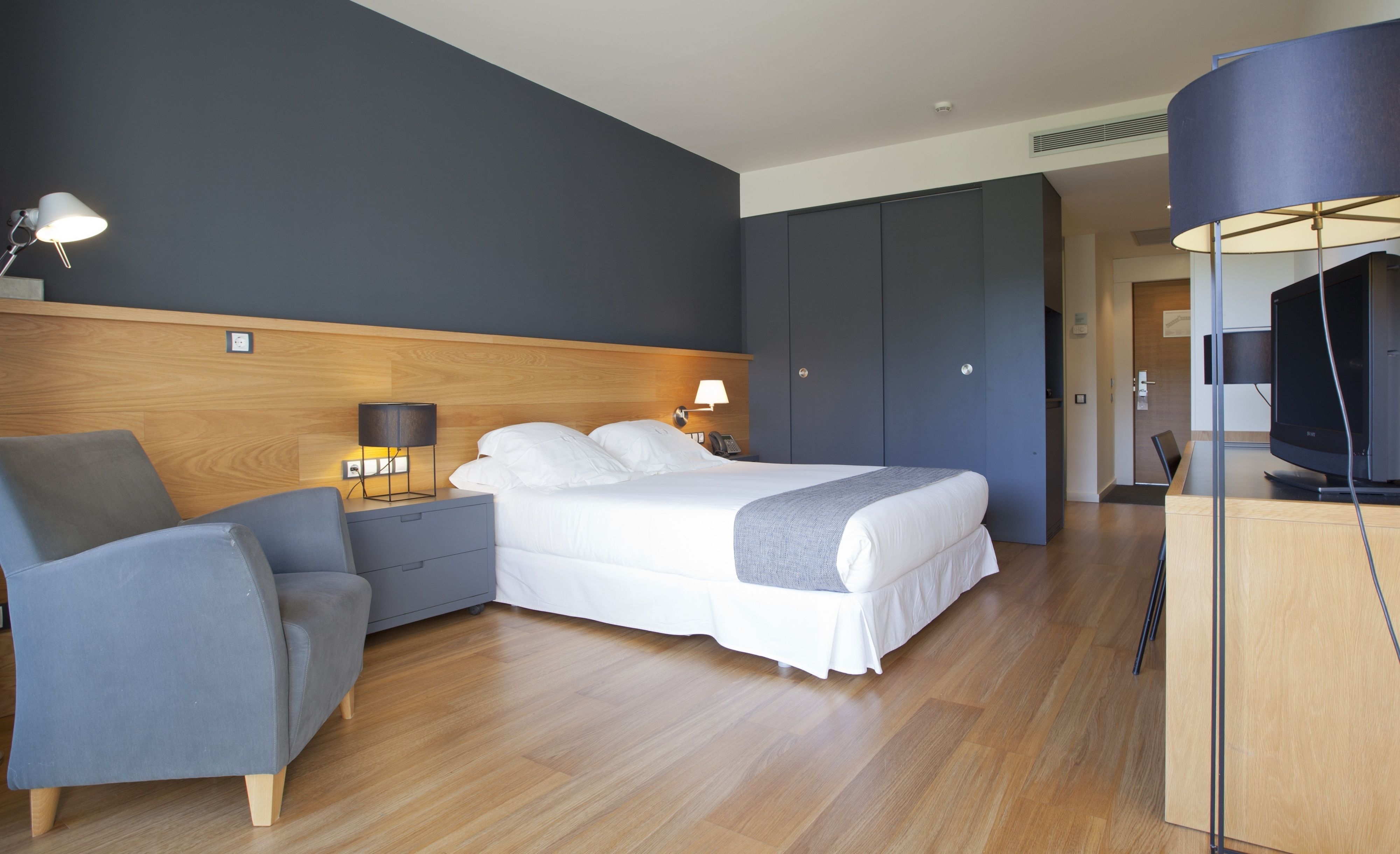 Quarto de hotel moderno com cama, mesas de cabeceira, escrivaninha, cadeira e televisão.