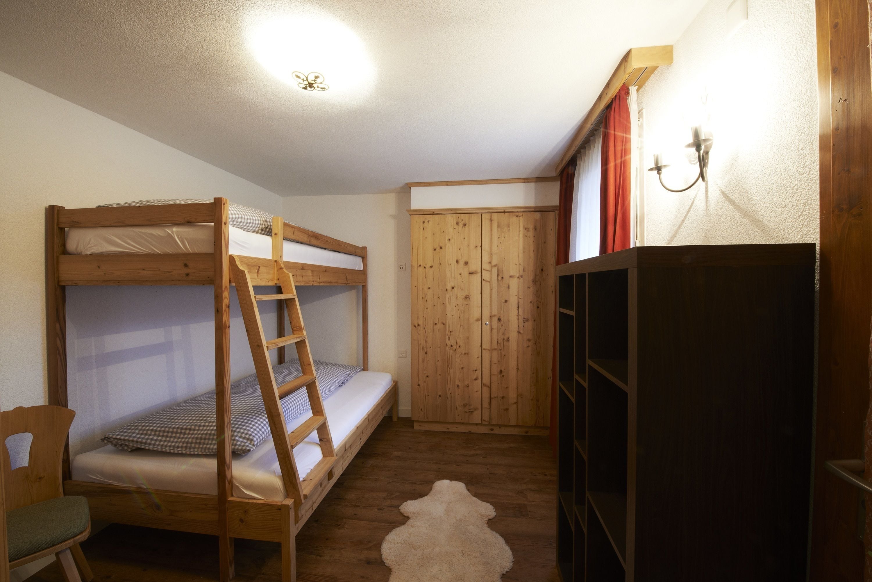 Uma cama beliche com escada, um guarda-roupa de madeira, uma mesa com cadeira e um tapete pequeno.