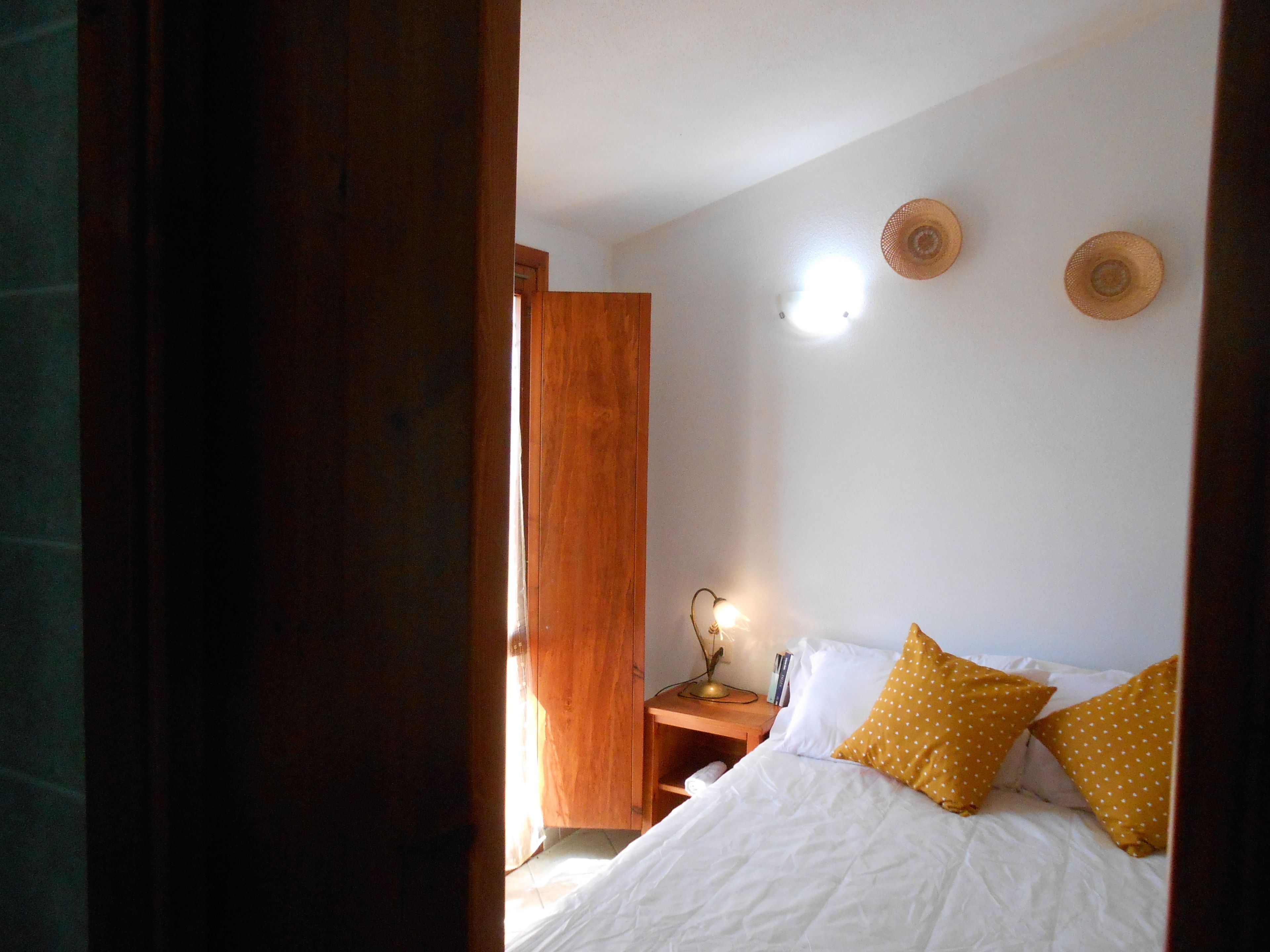 Quarto com guarda-roupa de madeira, mesa de cabeceira com abajur e dois chapéus decorativos na parede.