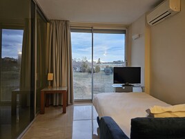 Quarto de hotel com uma cama, uma televisão, uma mesinha e vista da cidade por uma janela grande.
