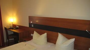 Quarto de hotel com cabeceira de madeira, duas camas com roupa de cama branca, uma escrivaninha com luminária e uma cadeira.