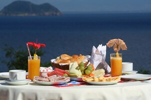 Mesa de café da manhã com diversos alimentos e bebidas, com vista para o oceano.