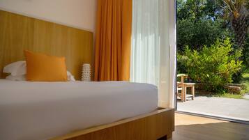 Quarto com cabeceira de madeira, cama grande com roupa de cama branca, almofada laranja e vista para um jardim através de uma porta de vidro.