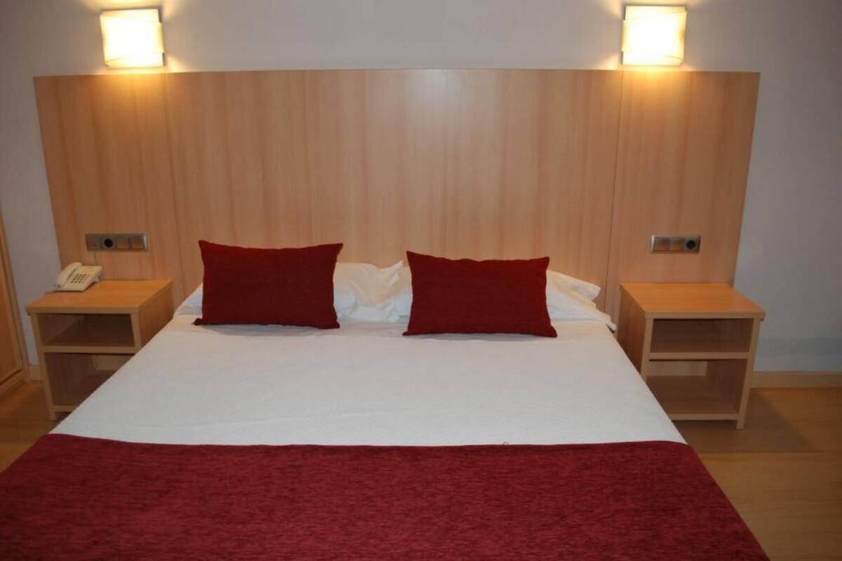 Quarto de hotel com cabeceira de madeira, duas almofadas vermelhas, colcha branca e mesas de cabeceira com abajures.