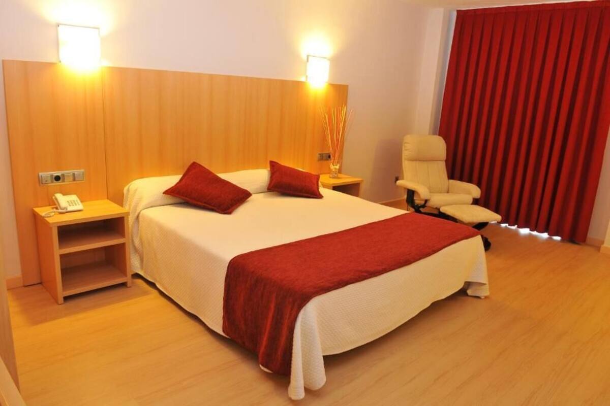 Quarto de hotel com uma cama grande, duas mesas de cabeceira, uma cadeira e uma cortina vermelha.