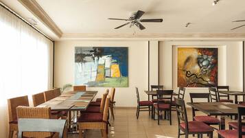Uma área de jantar com teto alto, ventiladores e duas grandes pinturas abstratas nas paredes.