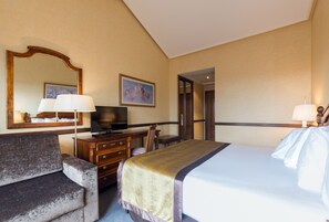 Quarto de hotel com cama, escrivaninha com televisão, penteadeira, cadeira, espelho e um quadro na parede.