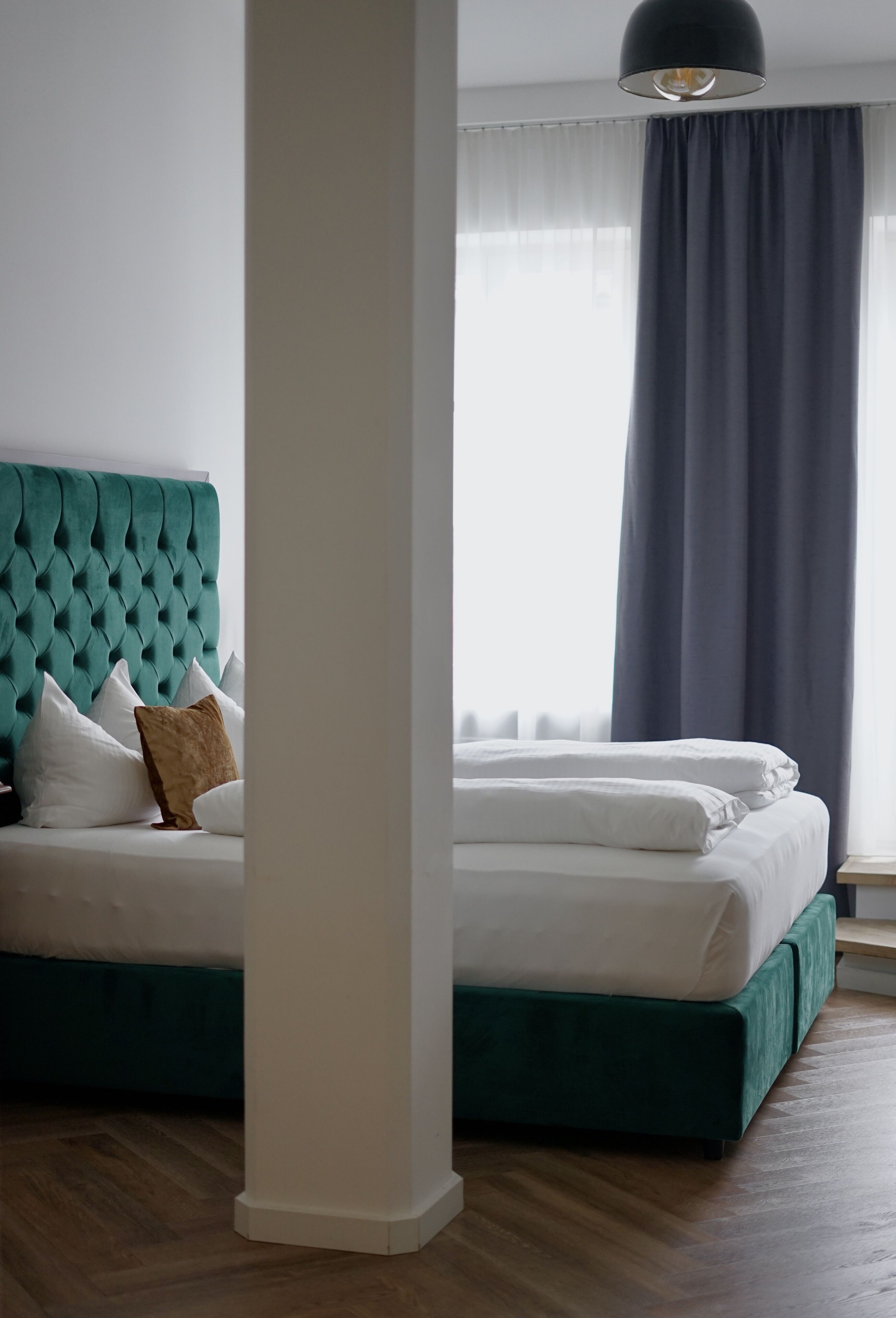 Uma cama bem arrumada com roupa de cama branca e cabeceira verde-água, uma janela grande com cortinas leves e piso de madeira.