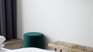 Quarto de hotel moderno com cama, um banquinho verde, mesa de cabeceira de madeira e TV montada na parede.