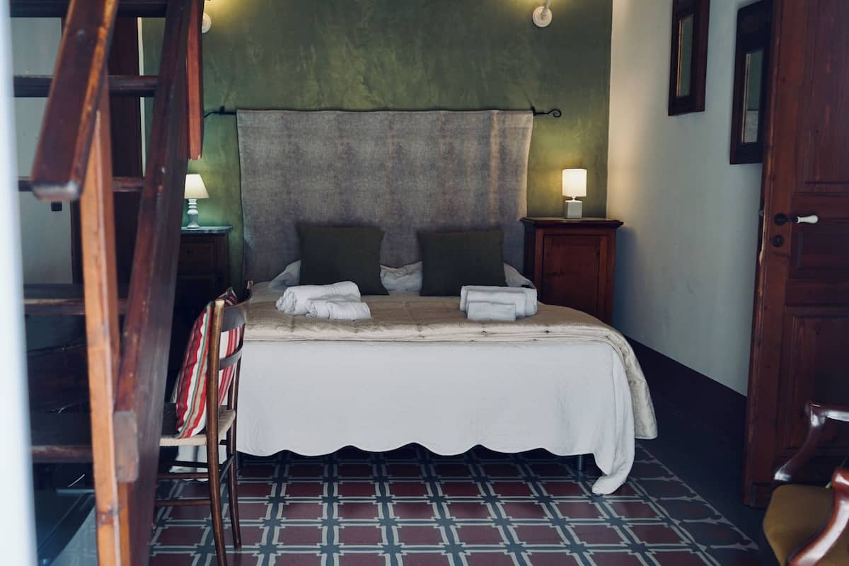 Cama bem arrumada com lençóis brancos e duas almofadas, uma cadeira de madeira, uma pequena mesa de cabeceira com abajur e piso estampado.