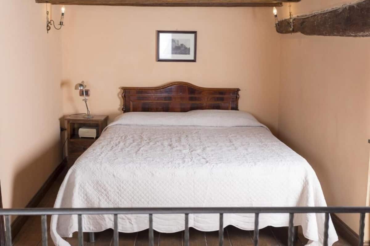 Quarto com cama de madeira, mesa de cabeceira, quadro na parede e vigamento de madeira no teto.