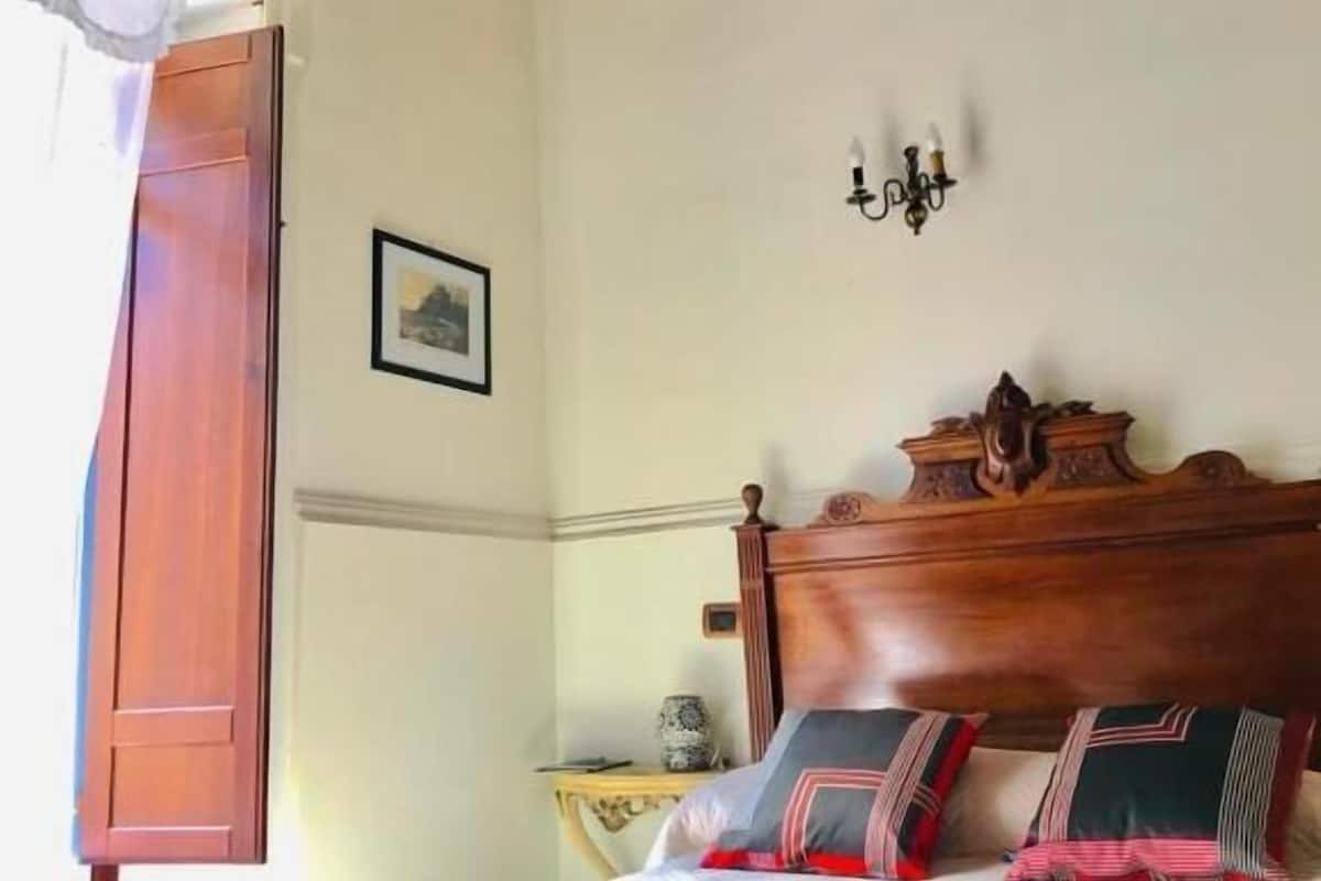 Quarto com cabeceira de madeira, cama com roupa de cama estampada, mesa de cabeceira com um vaso e luminária fixada na parede.