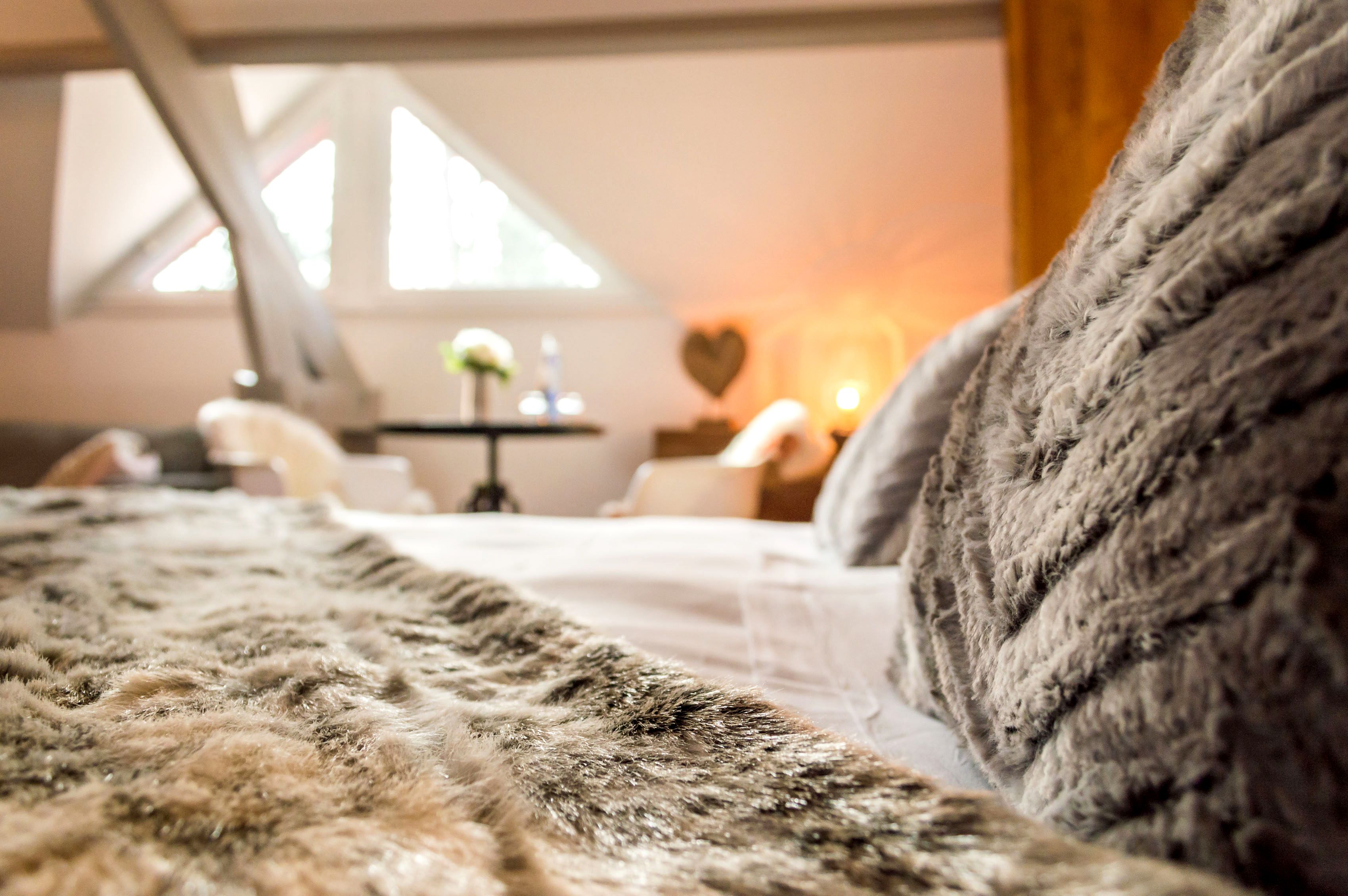 Um quarto com uma cama grande e fofa, uma mesa pequena com um vaso, e uma decoração em formato de coração na parede.