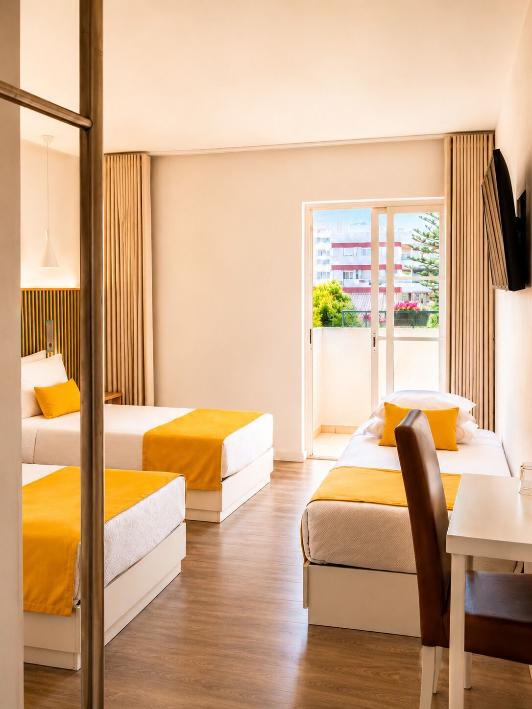 Quarto de hotel com duas camas, uma escrivaninha e uma cadeira. Há uma porta de vidro deslizante que dá para uma varanda com vista para edifícios e áreas verdes.