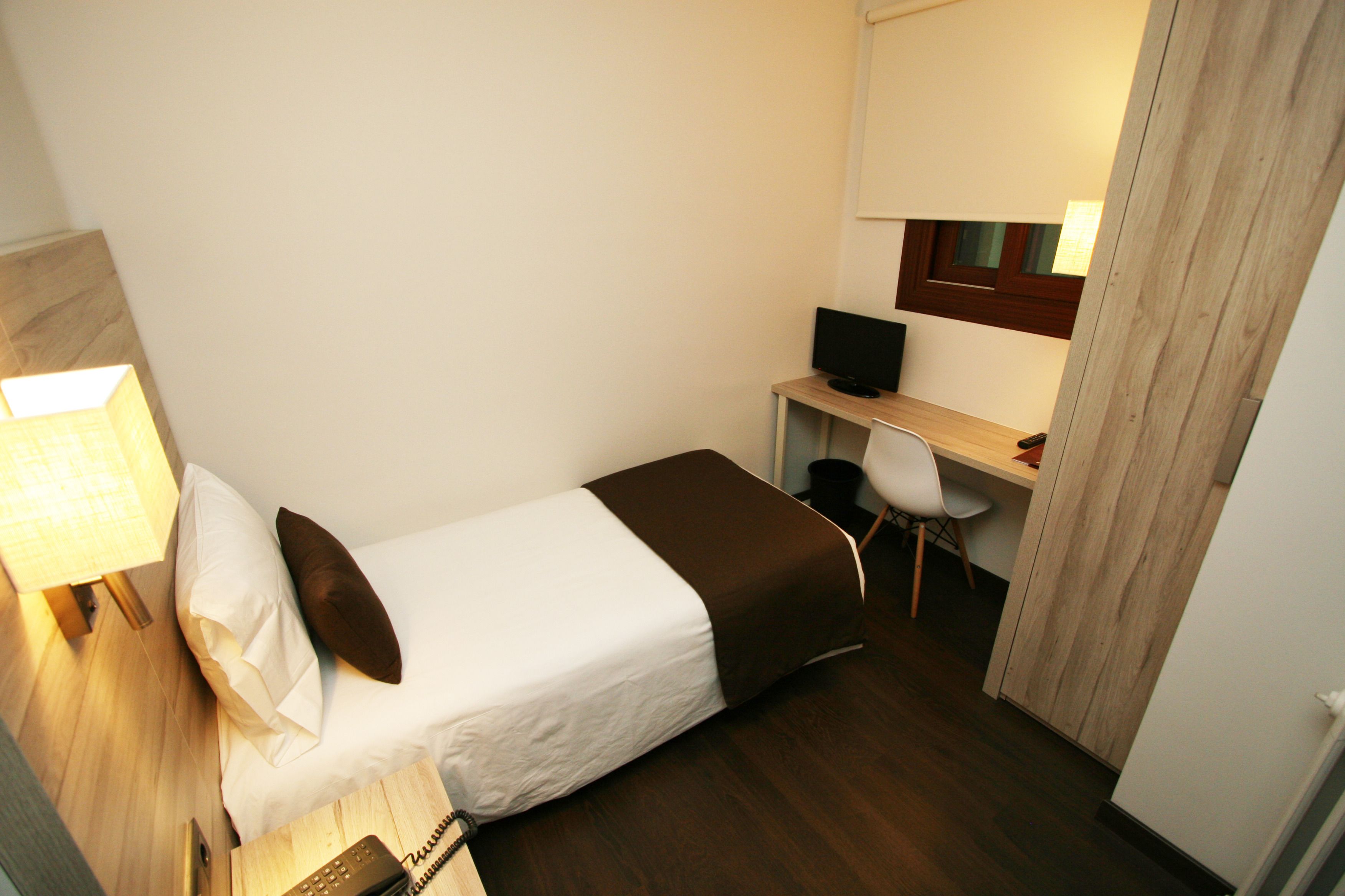 Quarto de hotel pequeno com uma cama, uma mesa, uma cadeira, um abajur e uma televisão.