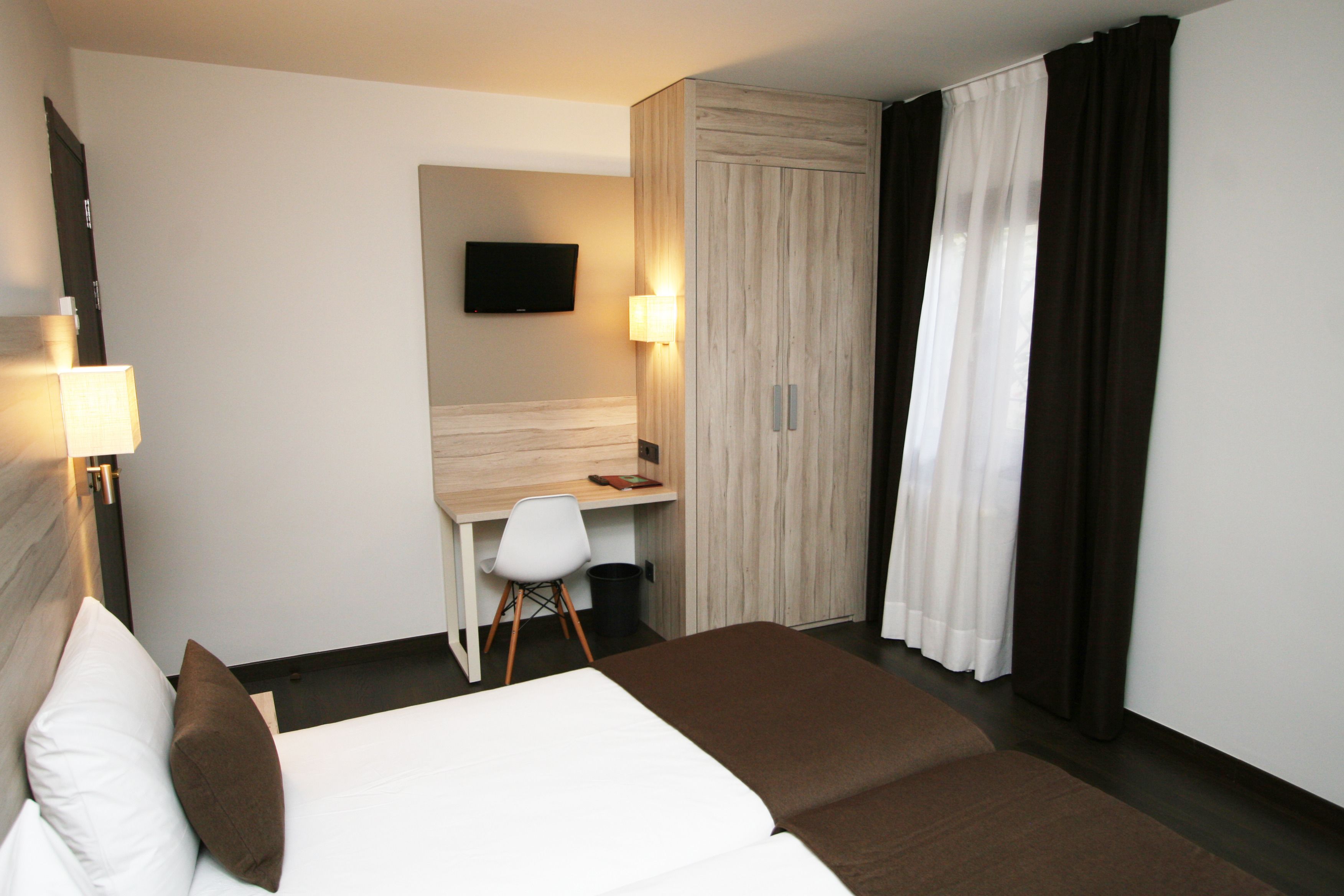 Quarto de hotel com uma cama, uma escrivaninha, uma cadeira, uma TV e cortinas.