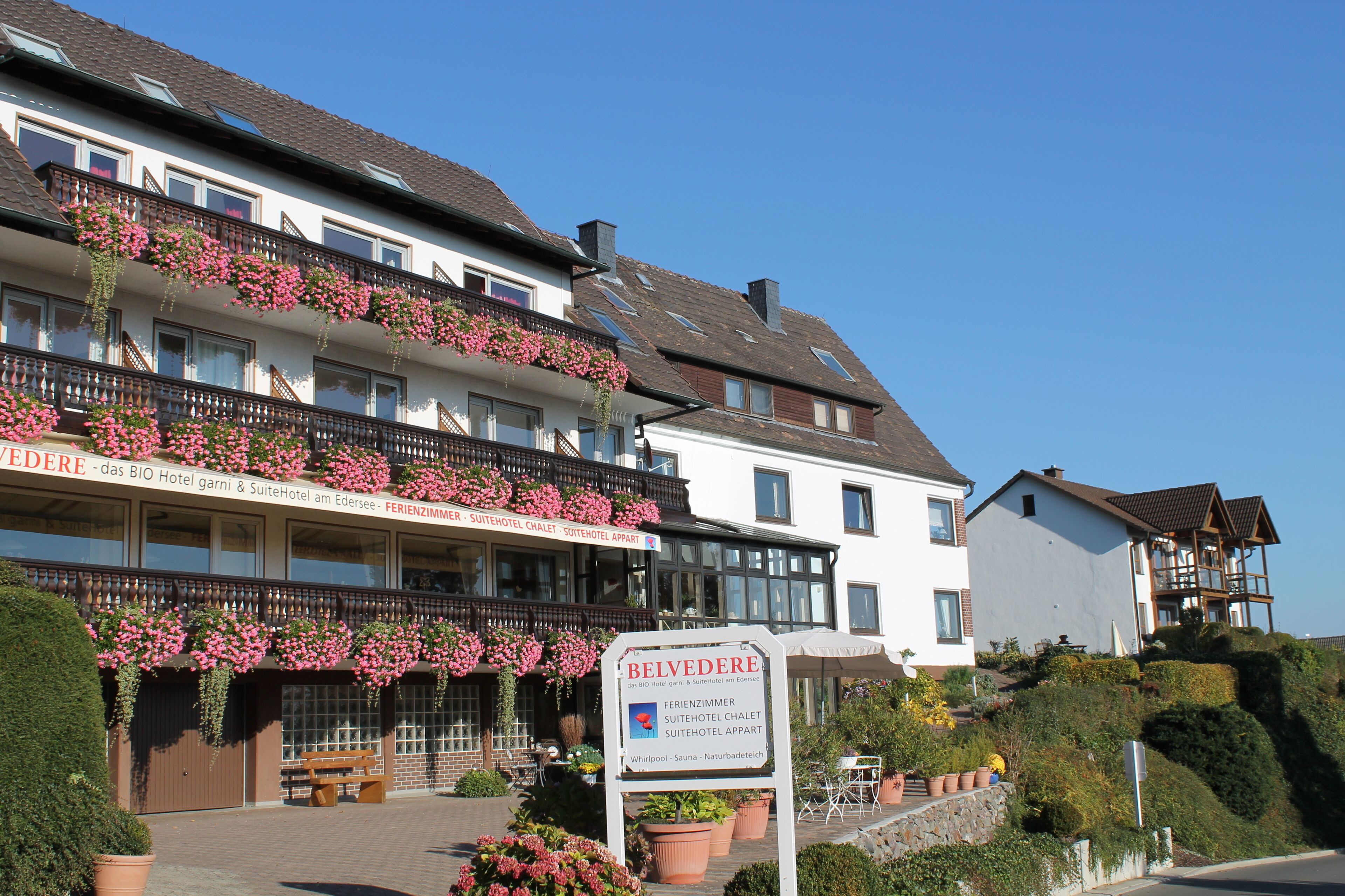 Um hotel com várias varandas decoradas com flores cor-de-rosa, uma placa indicando comodidades como sauna e restaurante, e vista para a colina circundante.