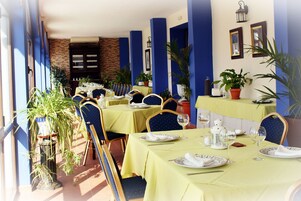 Restaurante com várias mesas preparadas para refeições, com cadeiras azuis e toalhas de mesa amarelas.