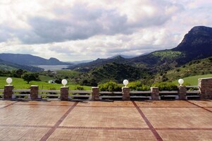 Um terraço pavimentado com grades de pedra e luminárias esféricas, com vista para uma paisagem montanhosa e um corpo d'água ao longe.