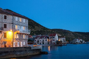 Uma vila costeira com edifícios de pedra, mar calmo e uma colina ao fundo.