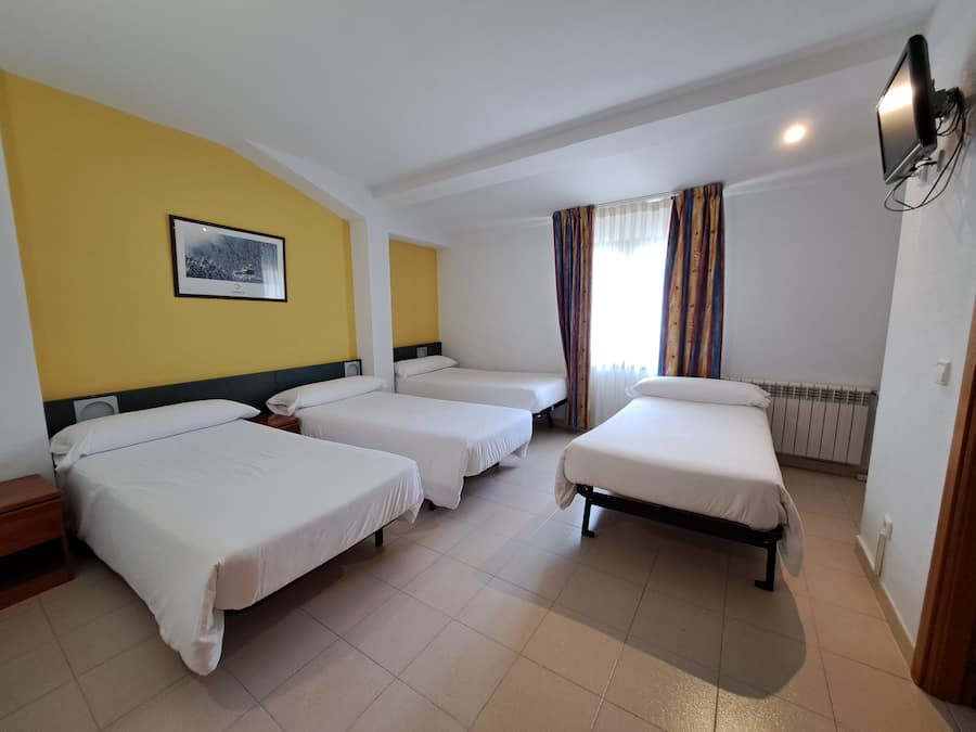 Quarto de hotel com três camas de solteiro, uma janela grande com cortinas, uma TV fixada na parede e um quadro emoldurado.