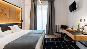 Quarto de hotel moderno com uma cama, uma escrivaninha, uma cadeira, uma televisão e uma janela com cortinas.