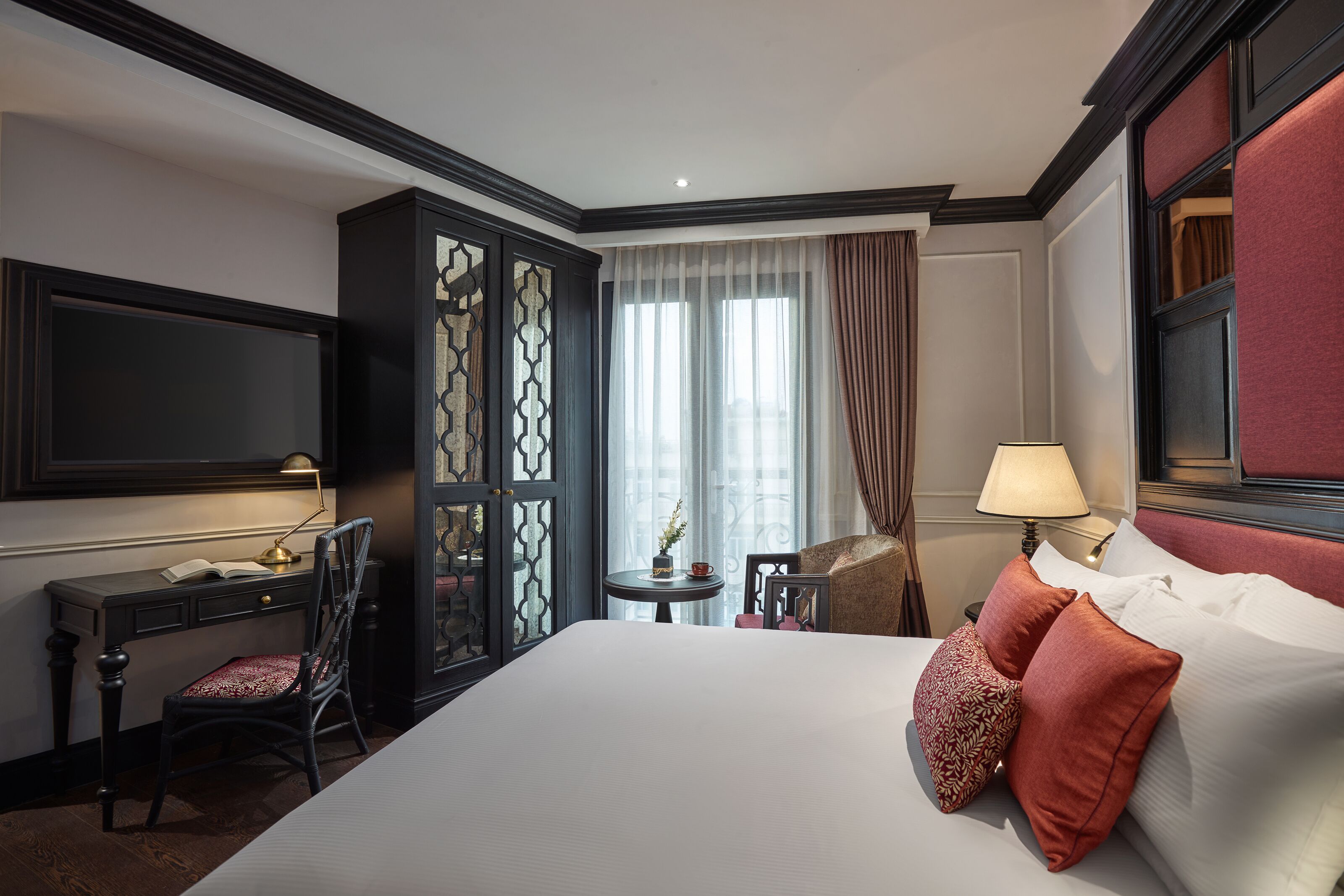 Quarto de hotel com uma cama grande, uma mesa com luminária, uma cadeira, uma televisão e uma janela com cortinas.