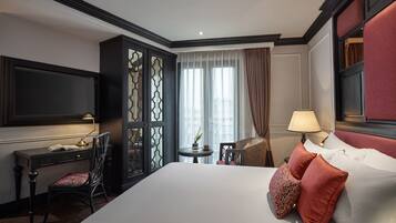 Quarto de hotel com uma cama grande, uma mesa com luminária, uma cadeira, uma televisão e uma janela com cortinas.