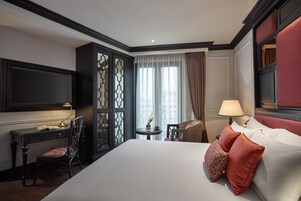 Quarto de hotel com uma cama grande, uma mesa com luminária, uma cadeira, uma televisão e uma janela com cortinas.