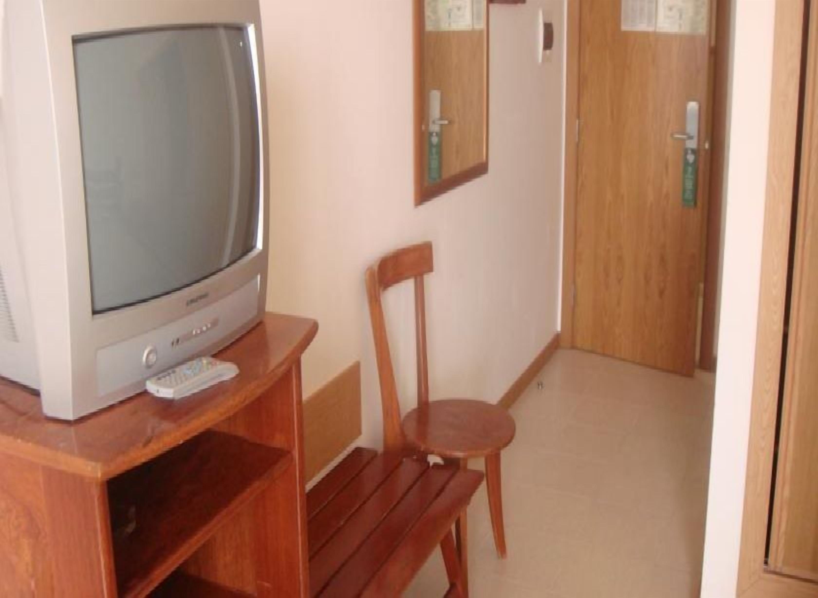 Quarto com cadeira de madeira, televisão sobre suporte de madeira e porta com espelho e interruptor de luz.