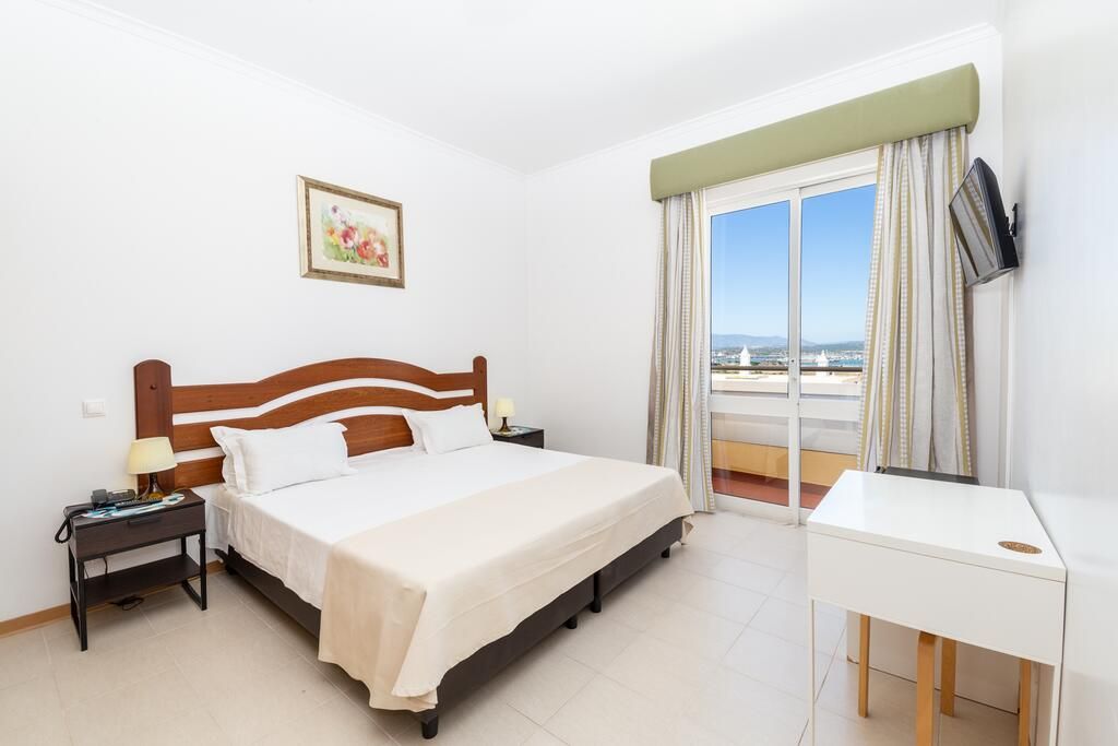 Quarto de hotel com cama, mesas de cabeceira, escrivaninha, janela com cortinas e um quadro na parede.