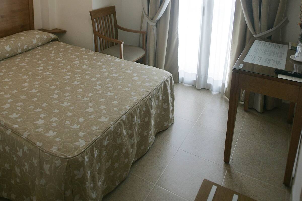 Quarto de hotel com cama, uma cadeira, escrivaninha com luminária e cortinas.