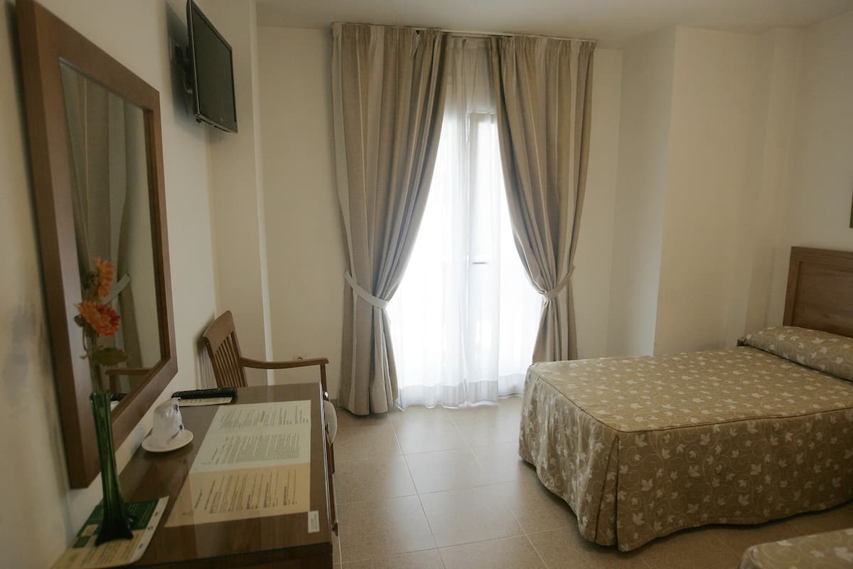 Quarto de hotel com uma cama, uma escrivaninha com cadeira, um espelho e uma janela com cortinas.