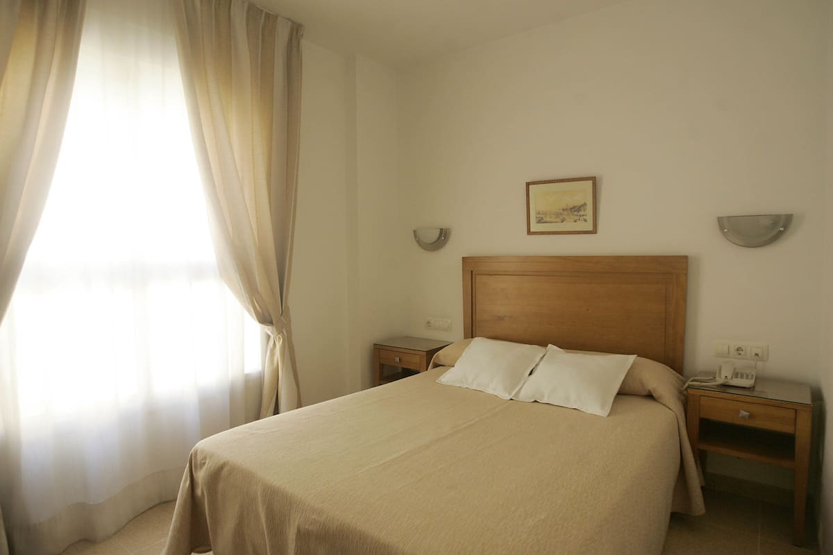 Quarto de hotel com cama, mesas de cabeceira, luminária e janela com cortinas.