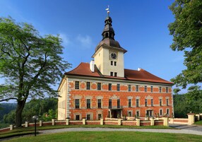 Um edifício grande e histórico com uma torre imponente e relógio, rodeado por vegetação exuberante e um jardim bem cuidado.