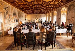 Uma sala de jantar formal com mesas postas para refeição, um grande mural em estilo afresco nas paredes e teto de madeira.