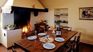 Uma sala de jantar com mesa de madeira posta para quatro pessoas, lareira e prateleiras com louças e um quadro.