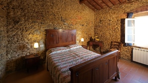 Quarto com parede de pedra, vigas de madeira no teto, uma cama com colcha, duas mesas de cabeceira com luminárias e uma janela com cortinas.