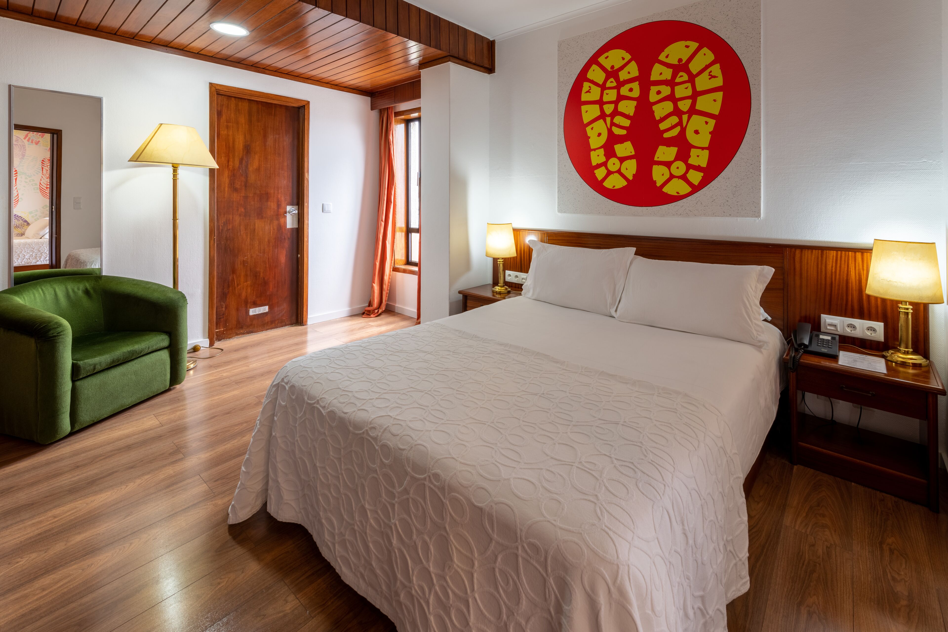 Quarto de hotel com uma cama grande, uma poltrona verde, piso de madeira e uma parede com uma obra de arte em vermelho e amarelo.