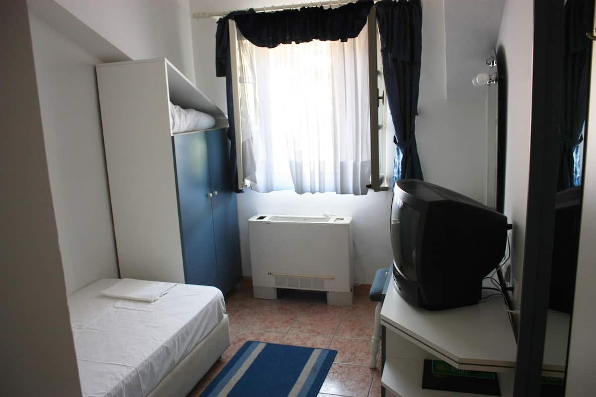Quarto pequeno e compacto, com uma cama, escrivaninha com computador, radiador e janela com cortinas.