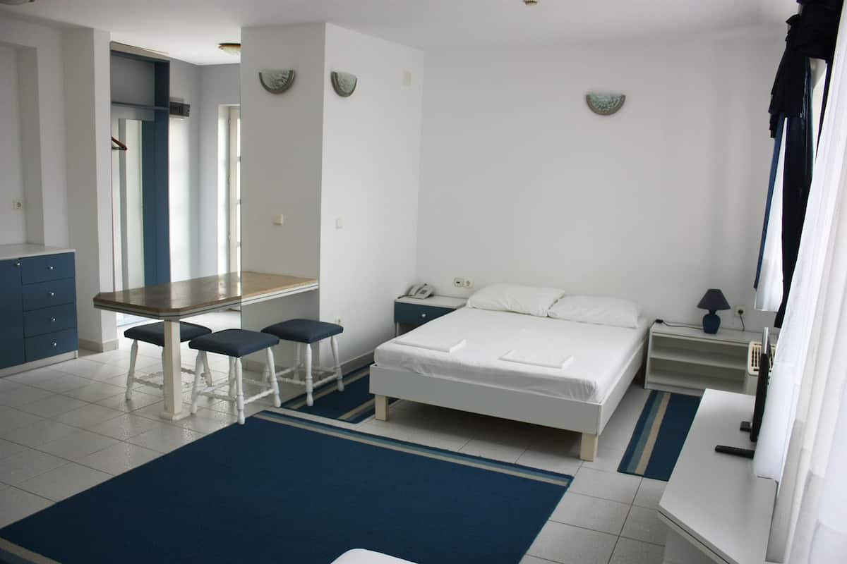 Quarto de hotel compacto com cama, mesa de cabeceira, criado-mudo e uma pequena escrivaninha com duas cadeiras.