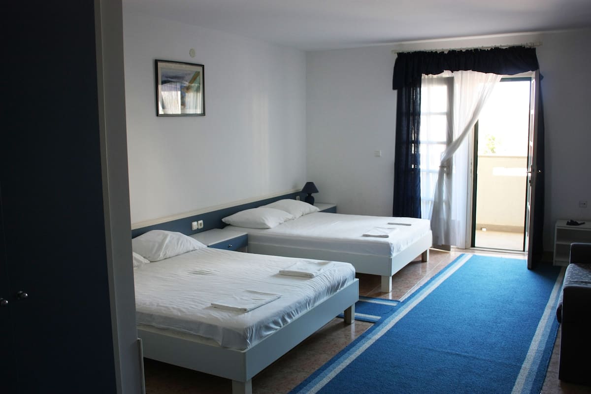 Quarto de hotel com duas camas de solteiro, uma pequena escrivaninha, uma cadeira, uma janela com cortinas e uma porta.