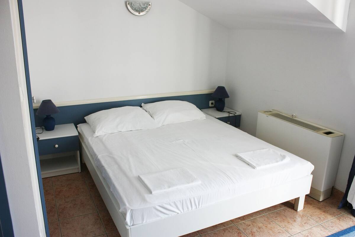 Uma cama bem arrumada com lençóis brancos e uma toalha dobrada, uma mesa de cabeceira com abajur e um aquecedor fixado na parede.