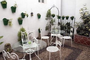 Mesa de vidro com cadeiras de metal branco, cercada por plantas em vasos contra uma parede branca.