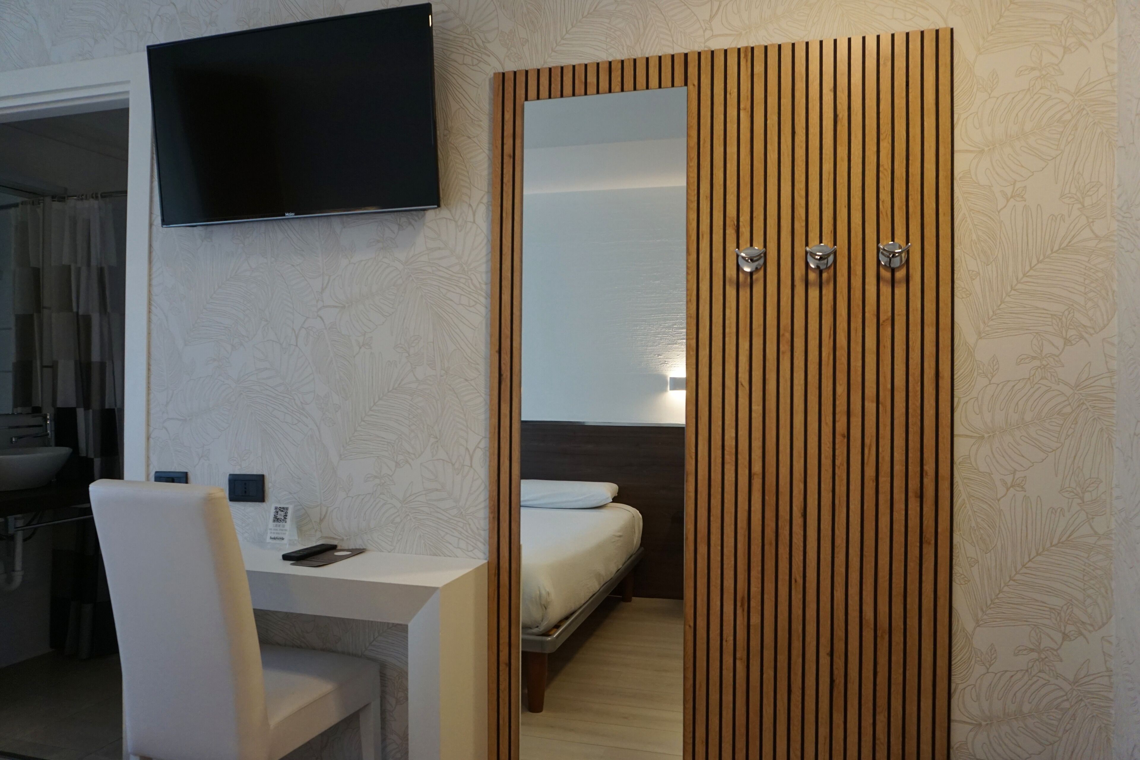 Quarto de hotel com guarda-roupa de madeira, uma cama, escrivaninha com cadeira, televisão na parede e banheiro com pia e espelho.