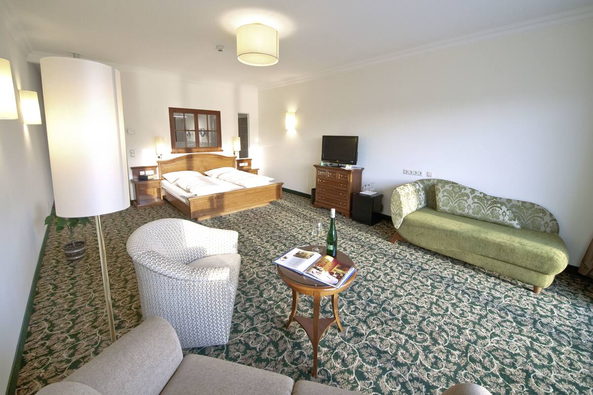 Quarto de hotel com cama, sofá verde, poltrona branca, cómoda de madeira, televisão e uma pequena mesa com uma revista e uma garrafa.